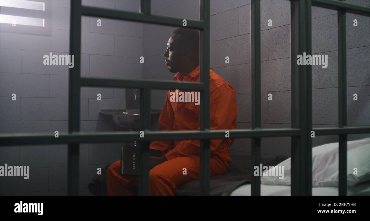 African American prisoner in orange uniform sits on the bed behind bars ...