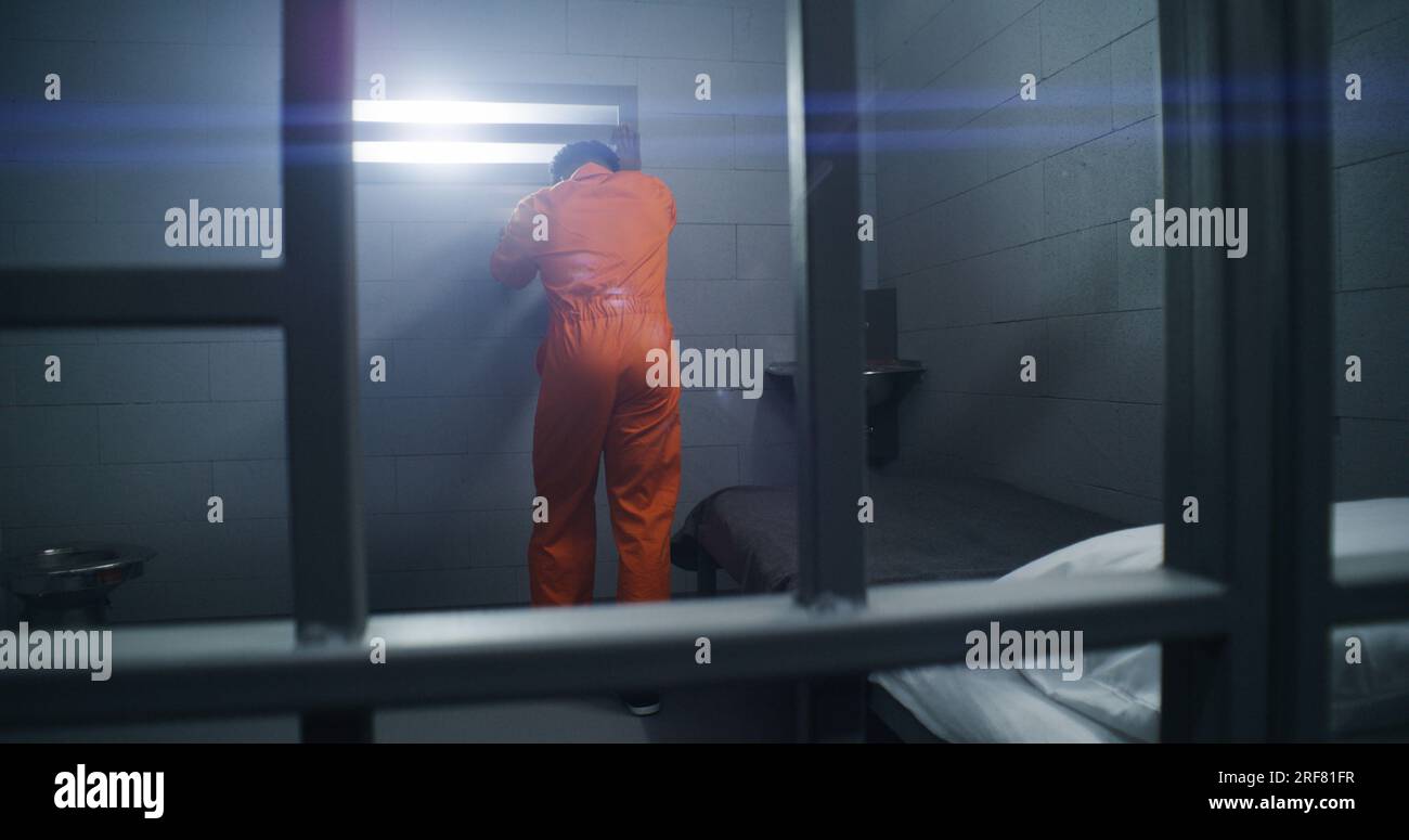 African American prisoner in orange uniform stands up from bed and leans on prison cell bars ...