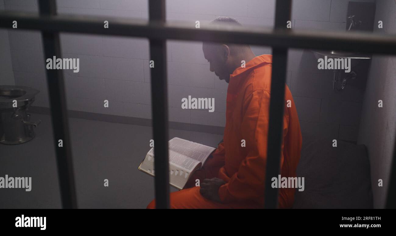 African American prisoner in orange uniform sits on bed behind bars ...