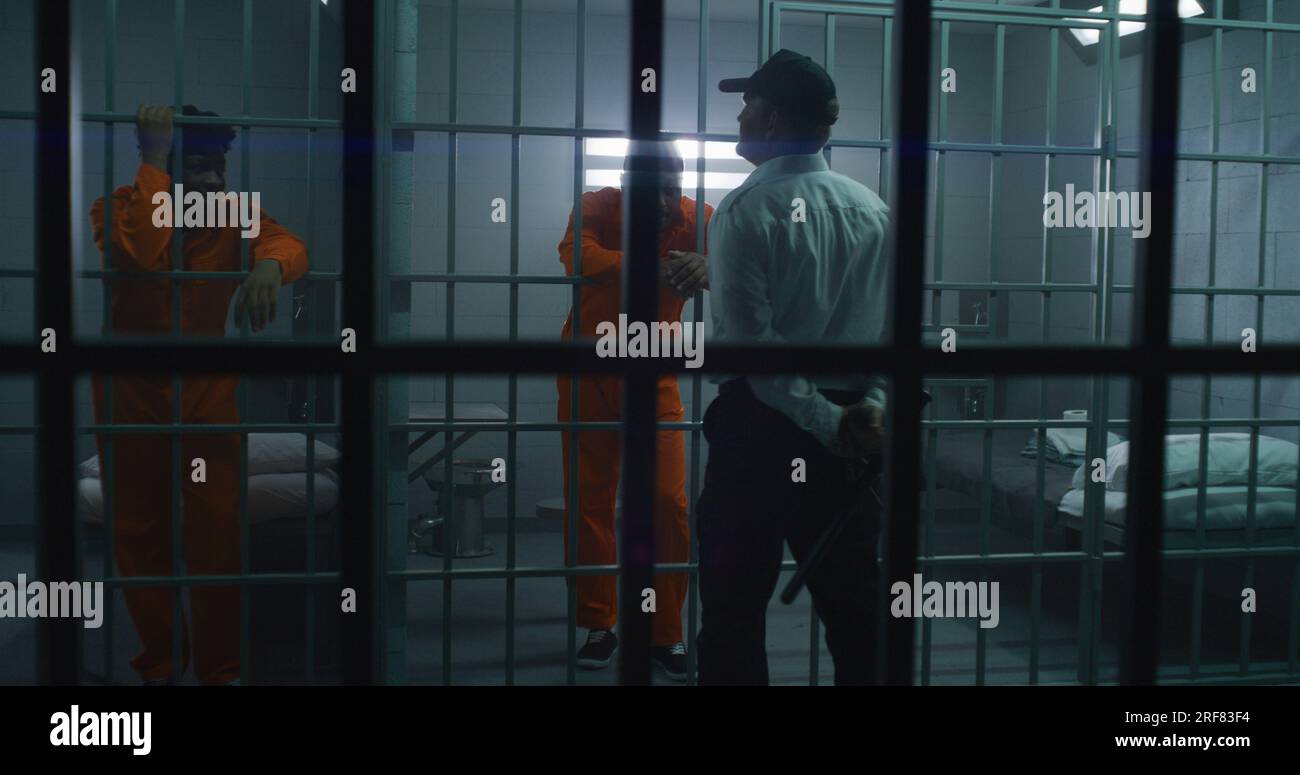 Jailer with police baton stands in front of jail cells and talks with ...