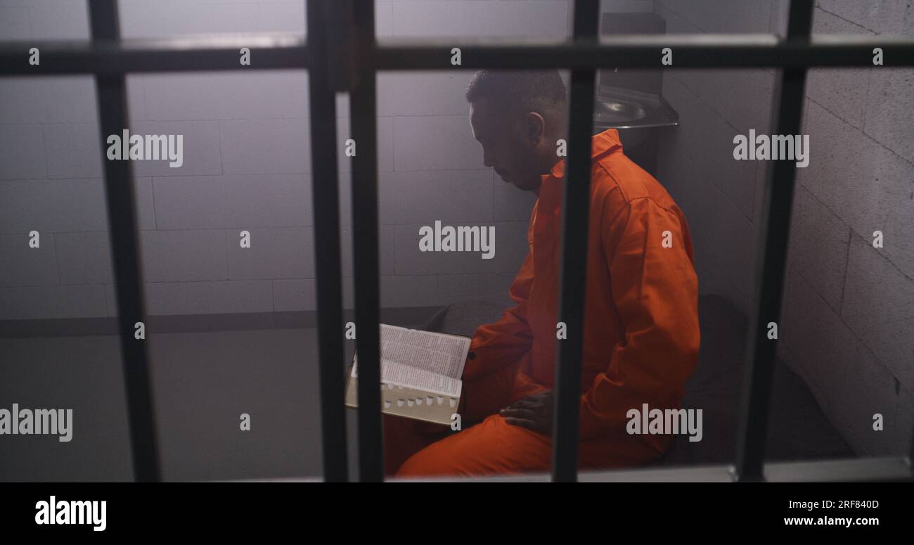 African American prisoner in orange uniform sits on bed behind bars ...