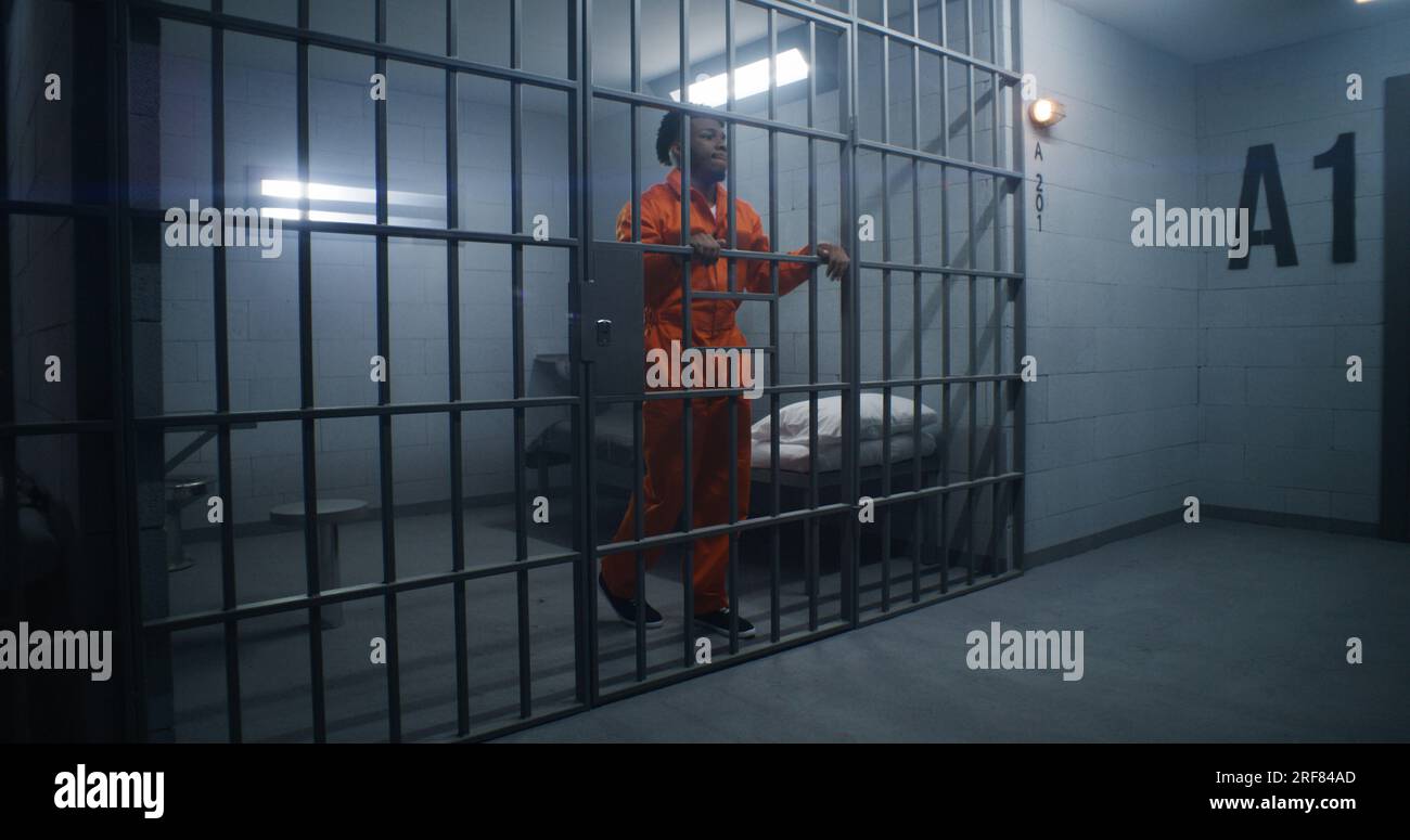 African American prisoner in orange uniform leans on jail cell bars and ...