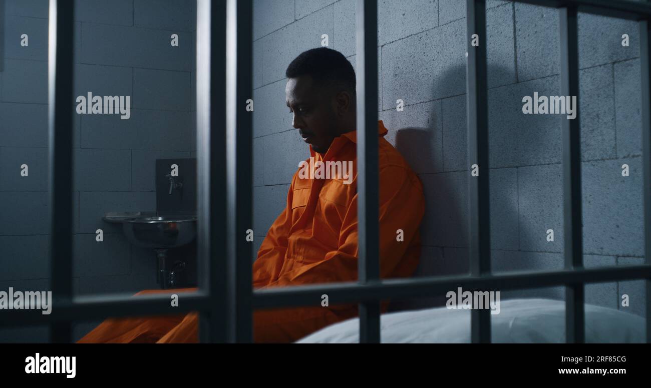 Guilty African American man in orange uniform sits on prison bed and ...