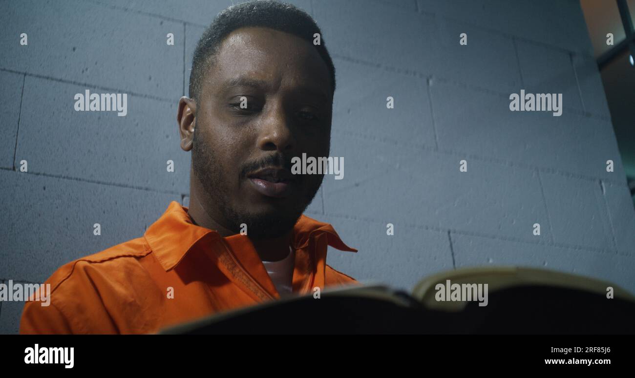 African American prisoner in orange uniform sits in prison cell, reads ...