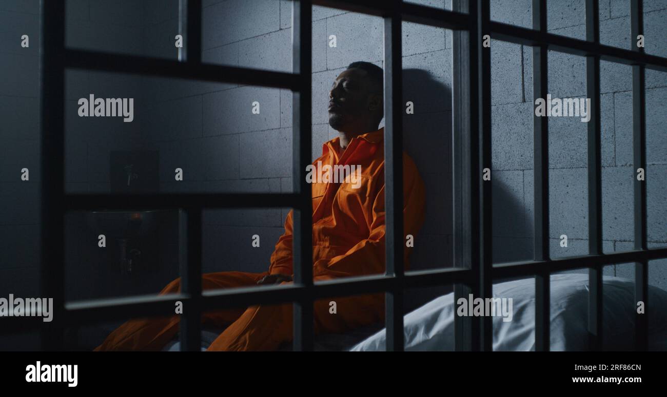 Guilty African American man in orange uniform sits on prison bed and ...
