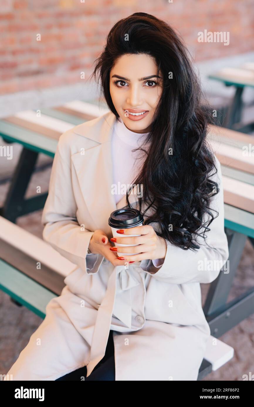 Adorable woman outdoors, white coat, sipping coffee, smiling for the ...