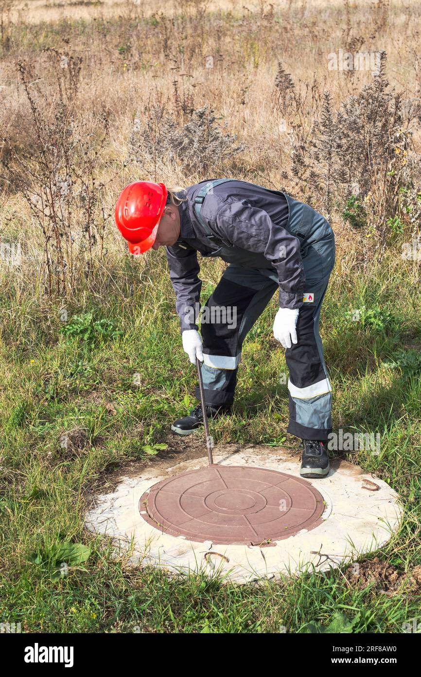 A worker in overalls and a helmet opens a manhole with a crowbar ...