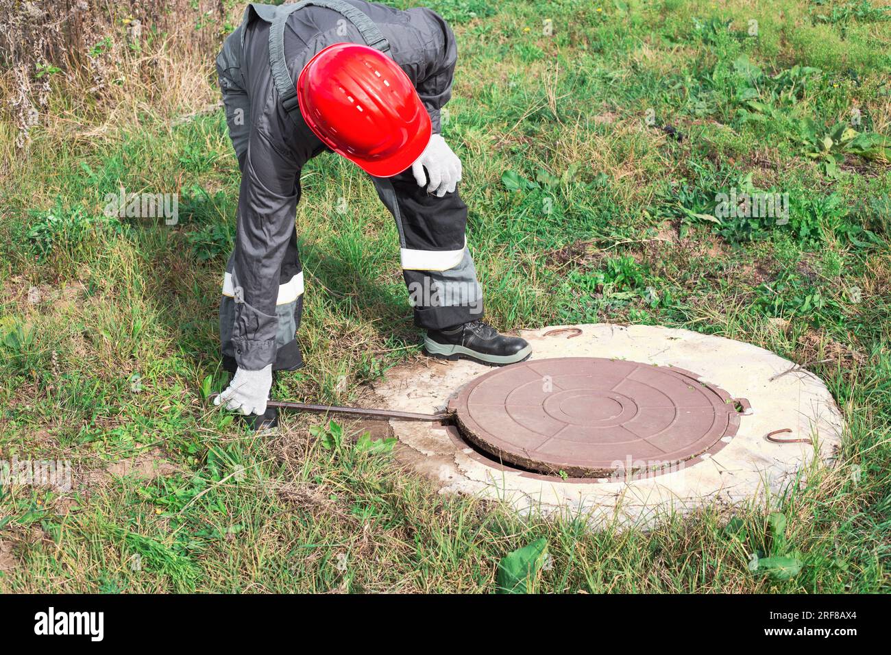 A worker in overalls and a helmet opens a manhole with a crowbar ...