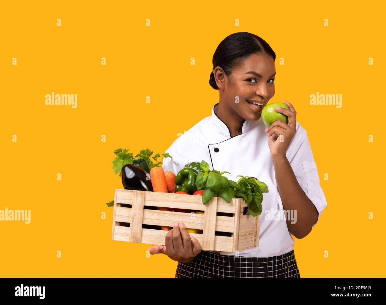 Black chef lady holding crate with vegetables aand fruits and biting ...