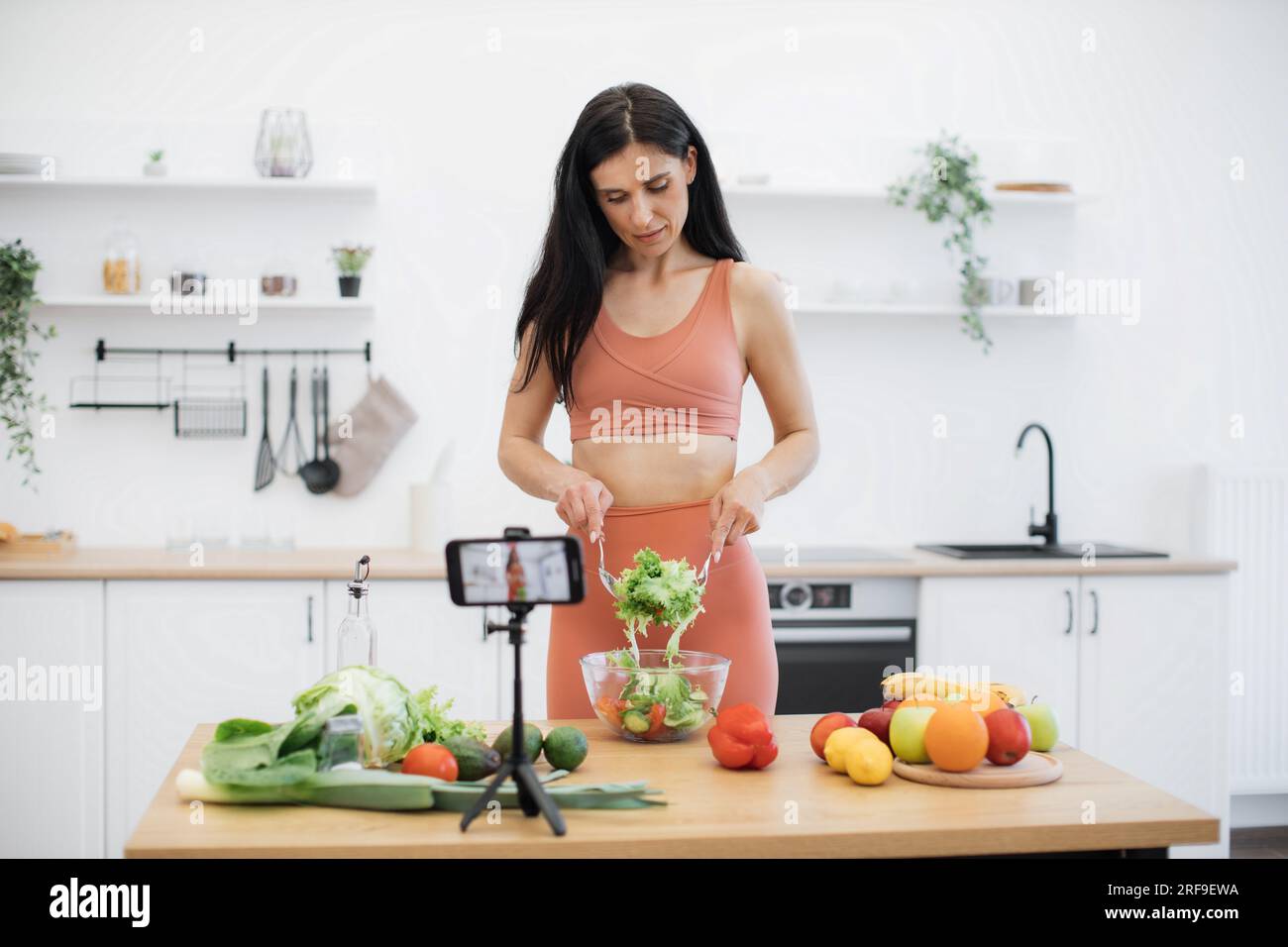 Happy woman in tight-fitting clothes tossing salad in glass bowl while ...