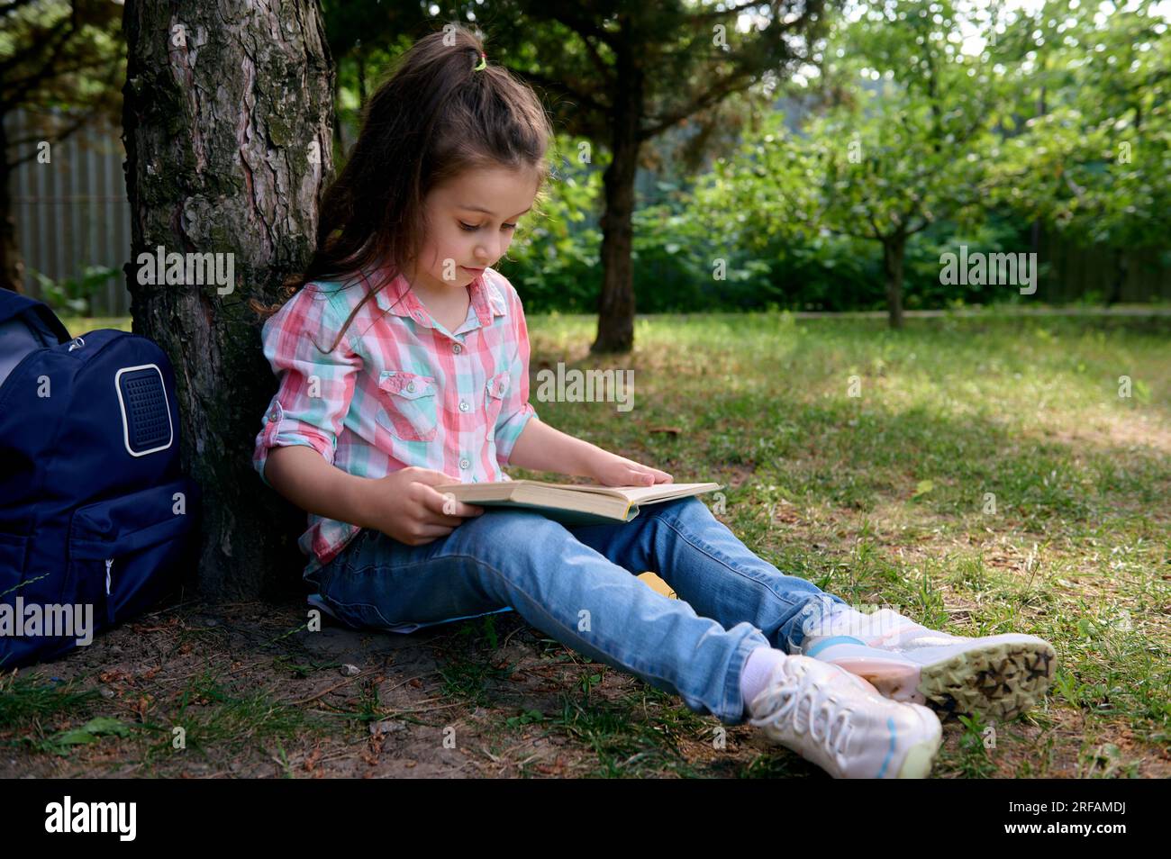 Little school girl reading a book in the park, sitting under a tree ...