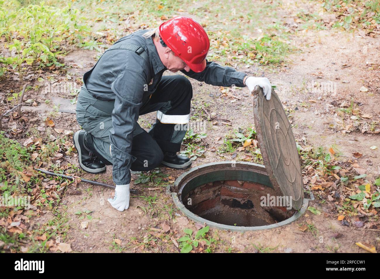 a man in overalls opened a sewer hatch and looks into a septic tank ...