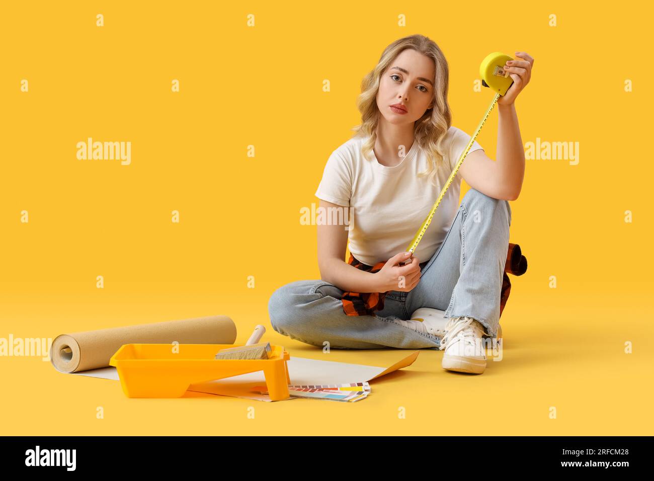 Upset young woman with measuring tape and wallpaper roll on yellow ...