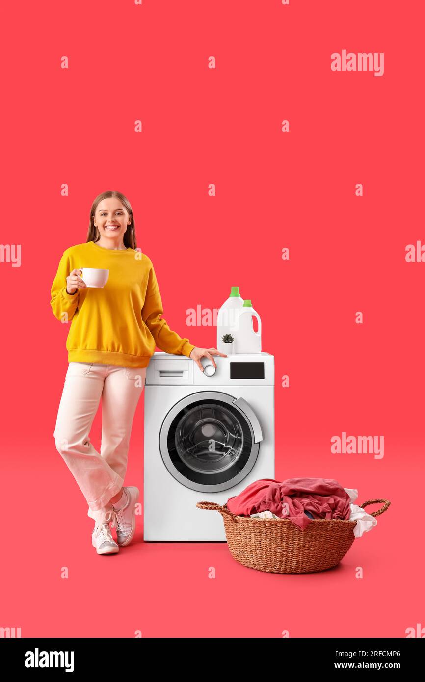 Pretty young woman with cup of tea setting up washing machine on red ...