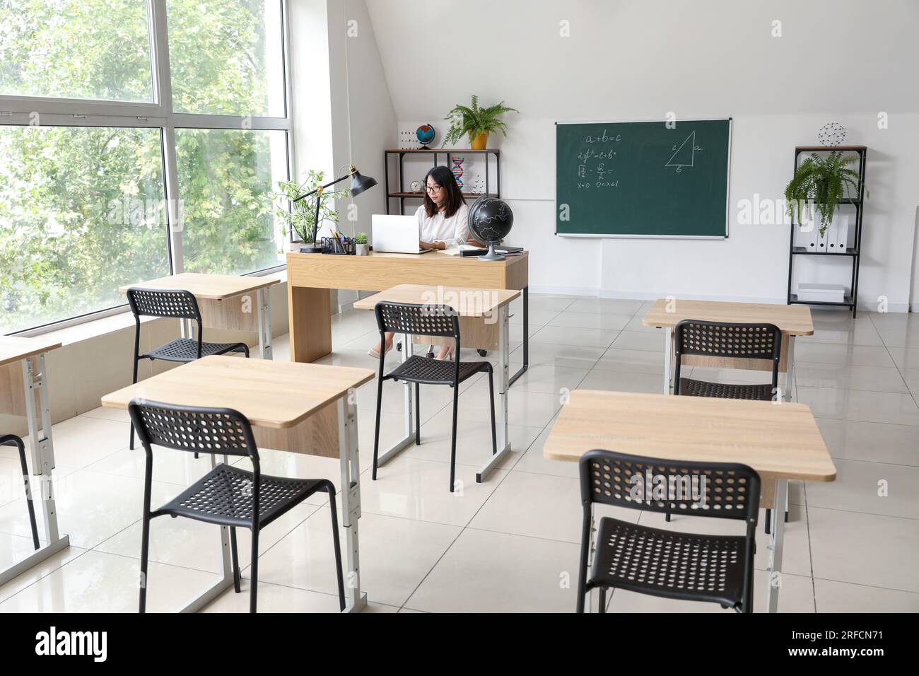 Female Asian teacher working with laptop at table in classroom Stock ...