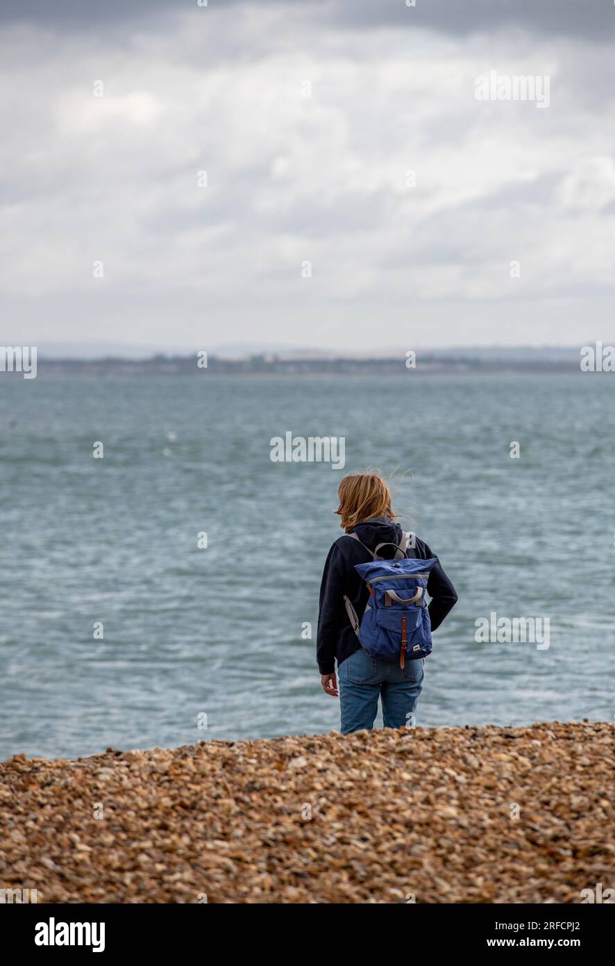 older woman standing on a shingle beach looking out to sea. middle aged ...