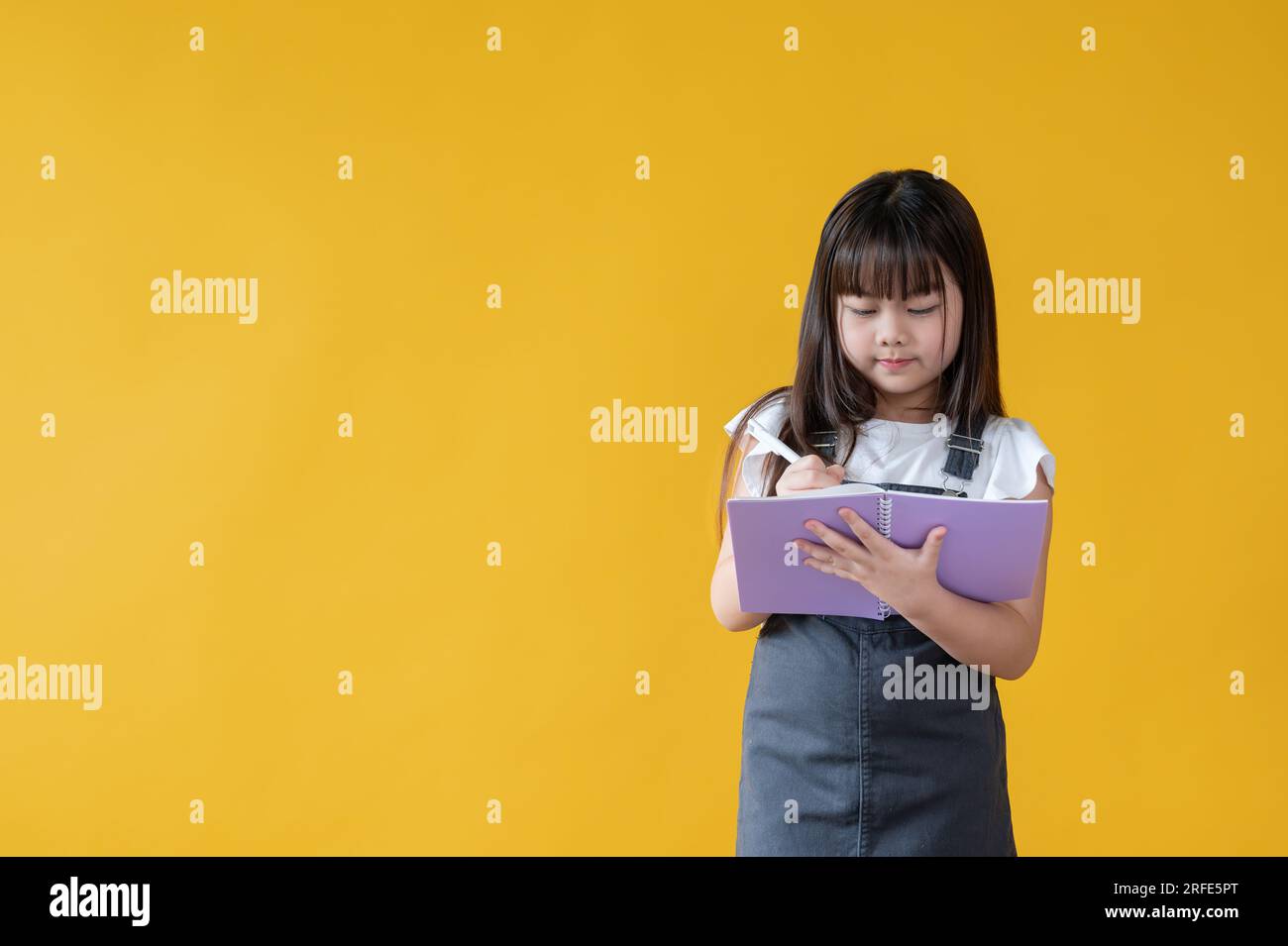 A cute little Asian girl is focusing on writing something in her book ...