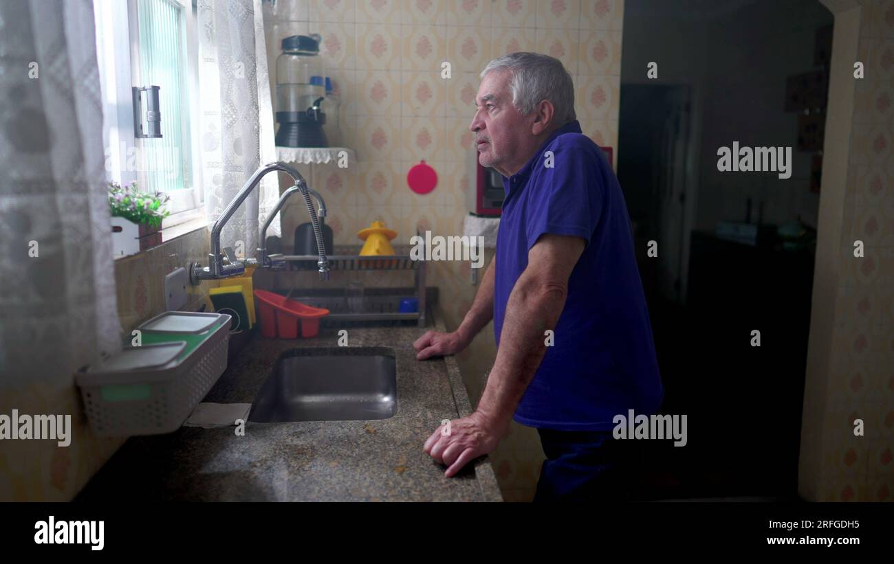 One pensive elderly man standing by kitchen sink staring out by window ...
