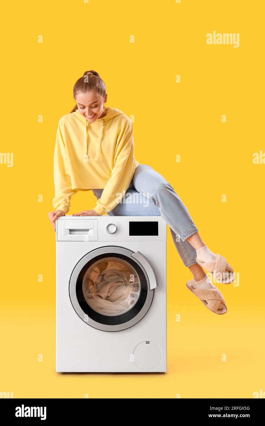 Pretty young woman sitting on washing machine against yellow background ...