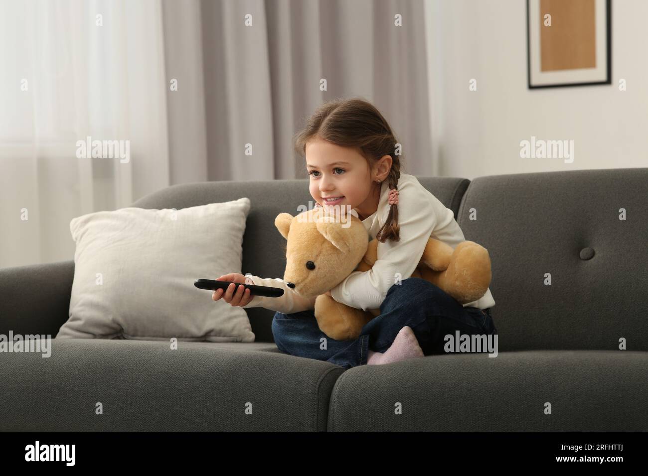 Little girl changing TV channels with remote control on sofa at home ...