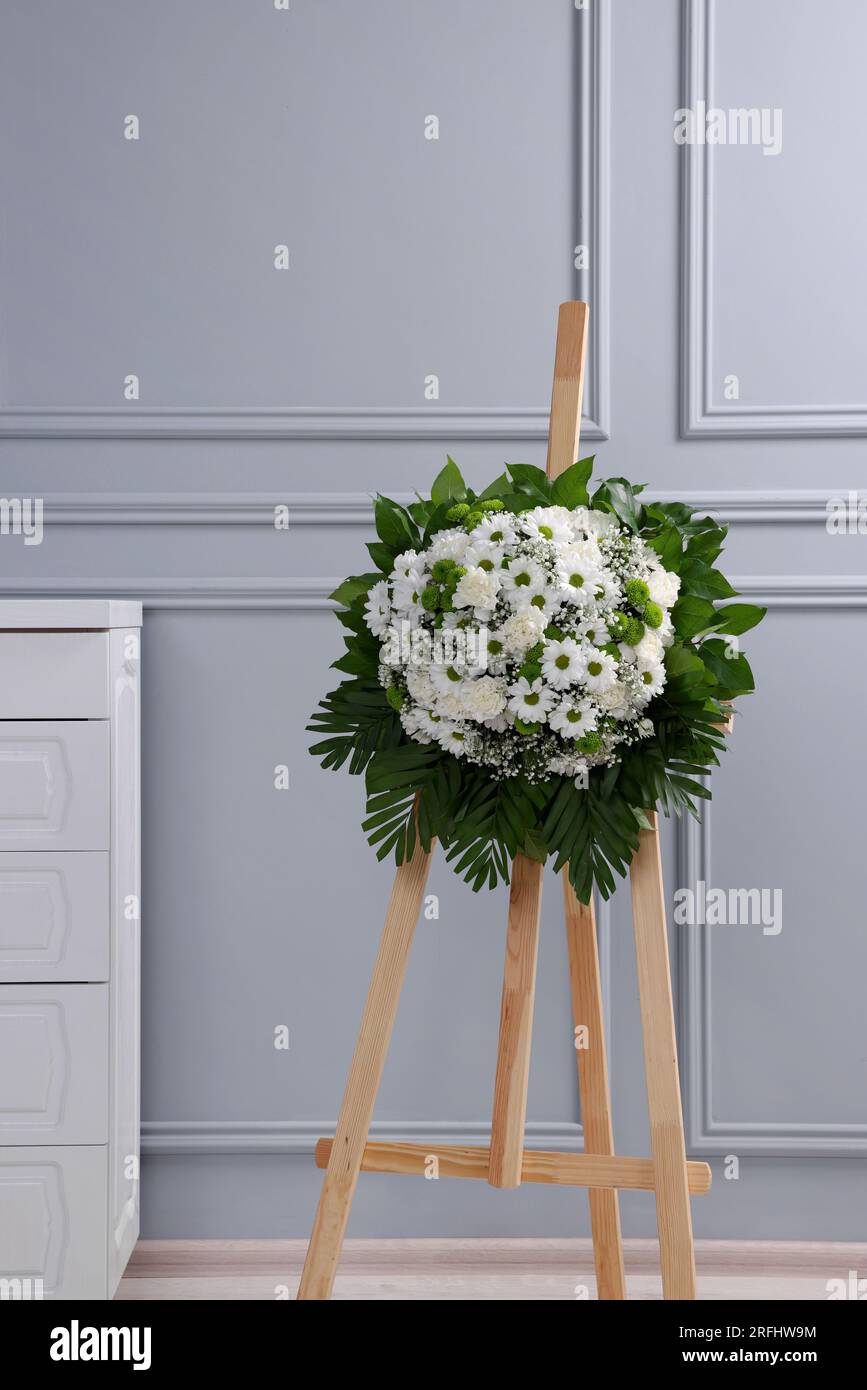 Funeral wreath of flowers on wooden stand near light grey wall indoors ...