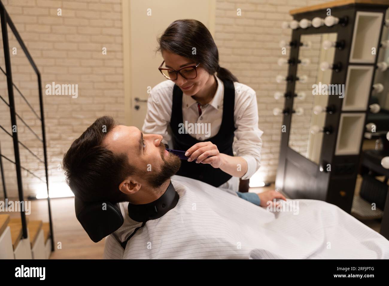 Female barber in glasses cuts the clients beard, the man sits in a comfortable chair Stock Photo ...