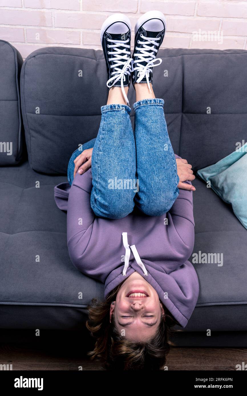 a teenage girl, lying on the couch upside down. a girl in blue jeans and hoodie sneakers relaxes ...