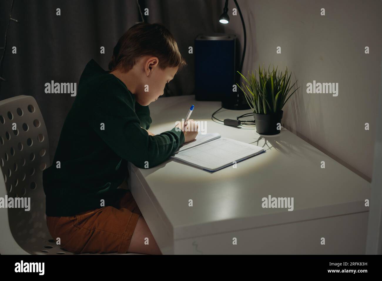 caucasian boy doing homework at home in dark room by the light of lamp ...