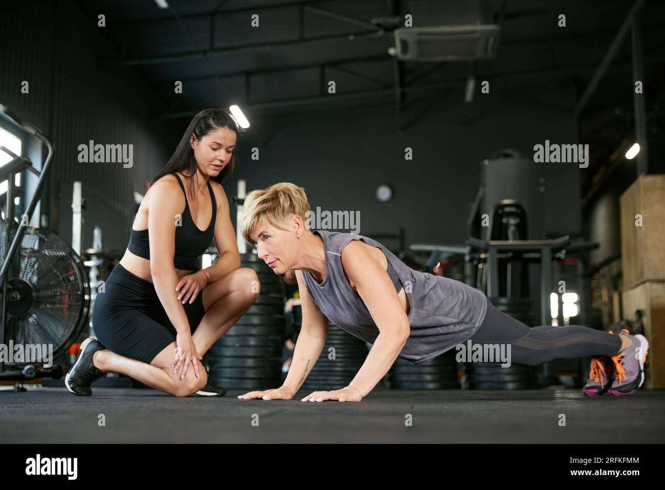Sportive mature woman training in gym with female fitness trainer, doing push ups exercises ...