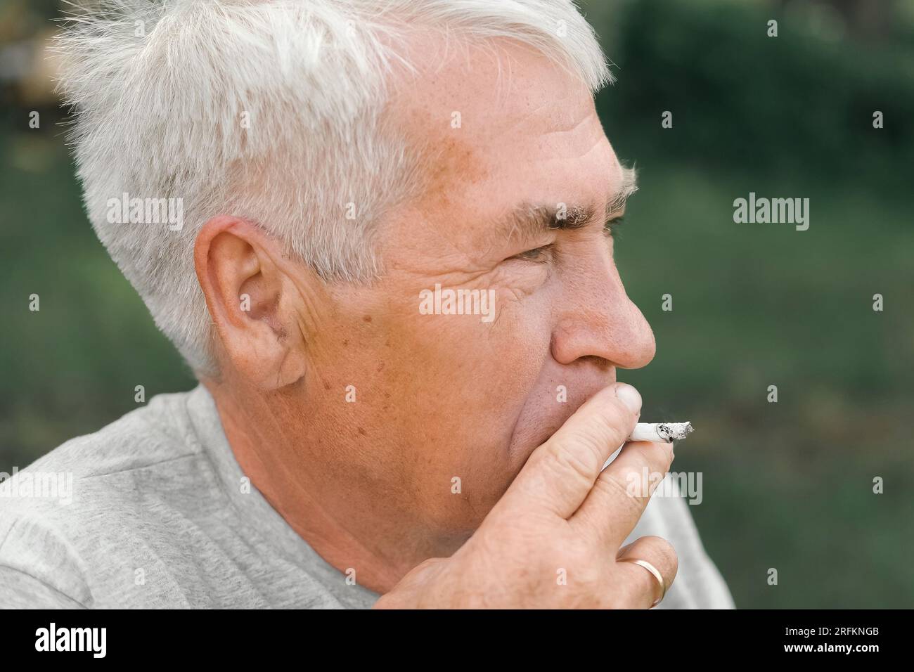 Senior man lighting up a cigarette outdoors against green nature ...