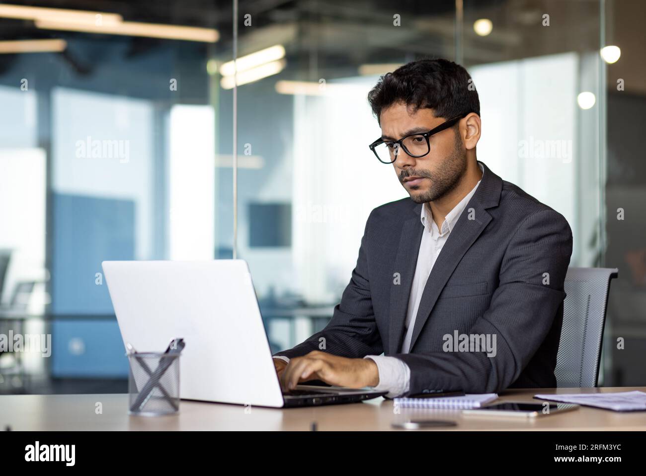 Young serious and focused businessman at workplace typing on computer, Arab man in business suit ...