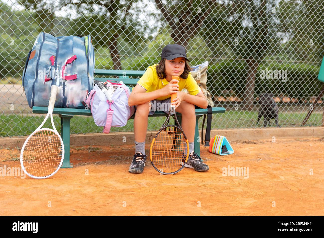 A boy sitting on a bench with sports bafs by the tennis court and ...