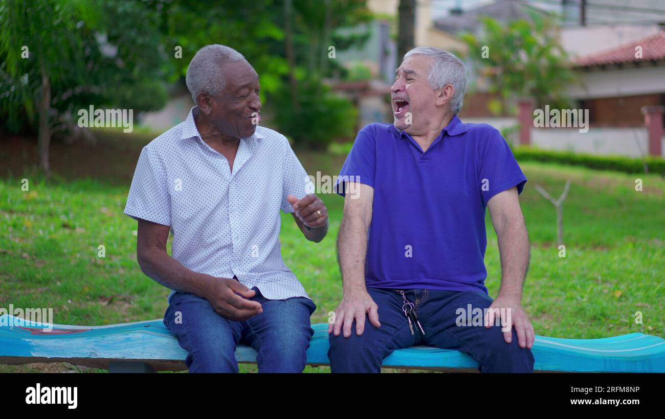 Two diverse senior talking and laughing sitting on park bench in Brazil ...