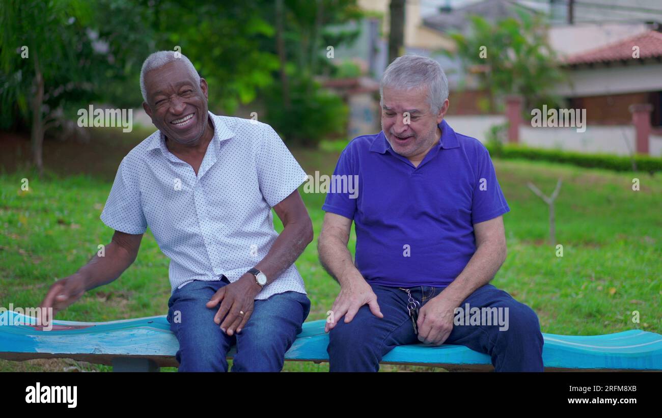 Two diverse senior talking and laughing sitting on park bench in Brazil ...