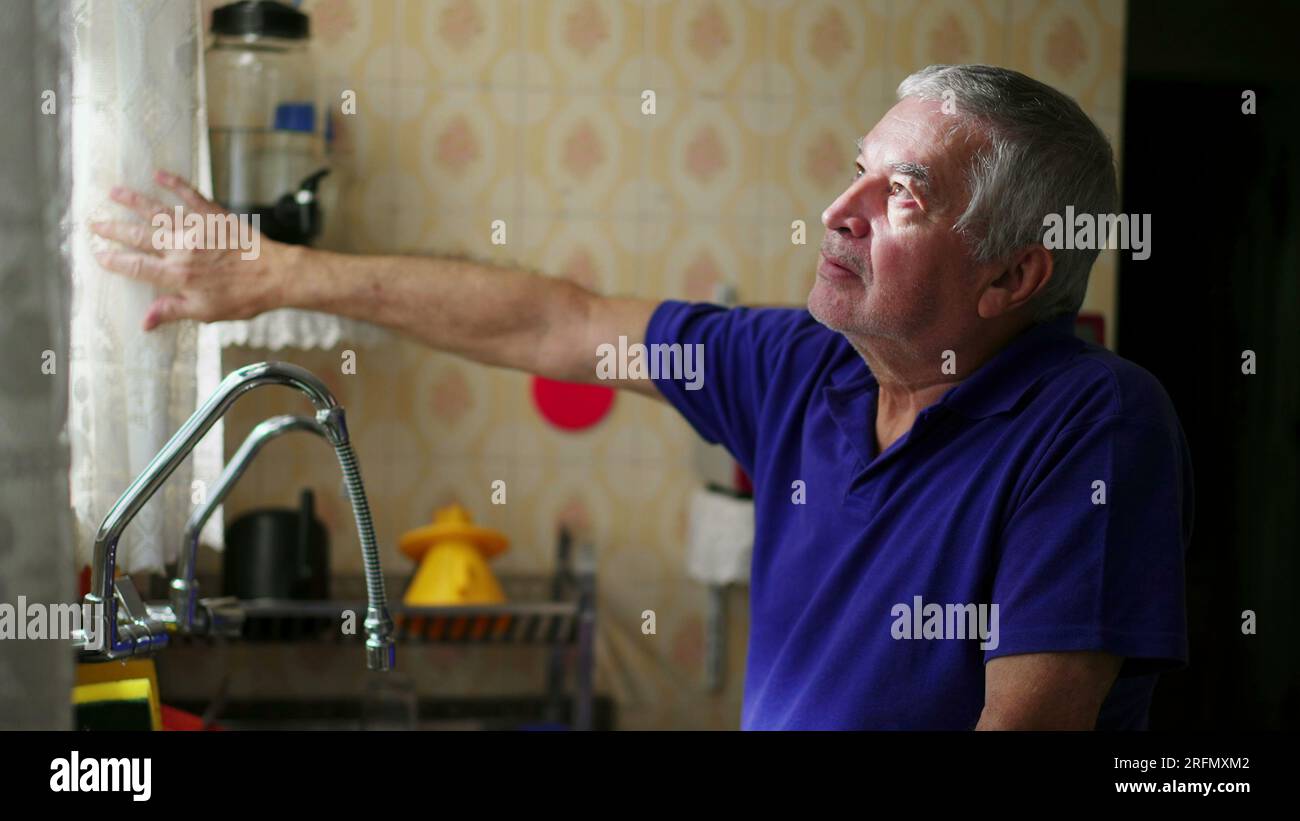 Senior man standing by kitchen window peeking out through curtain ...