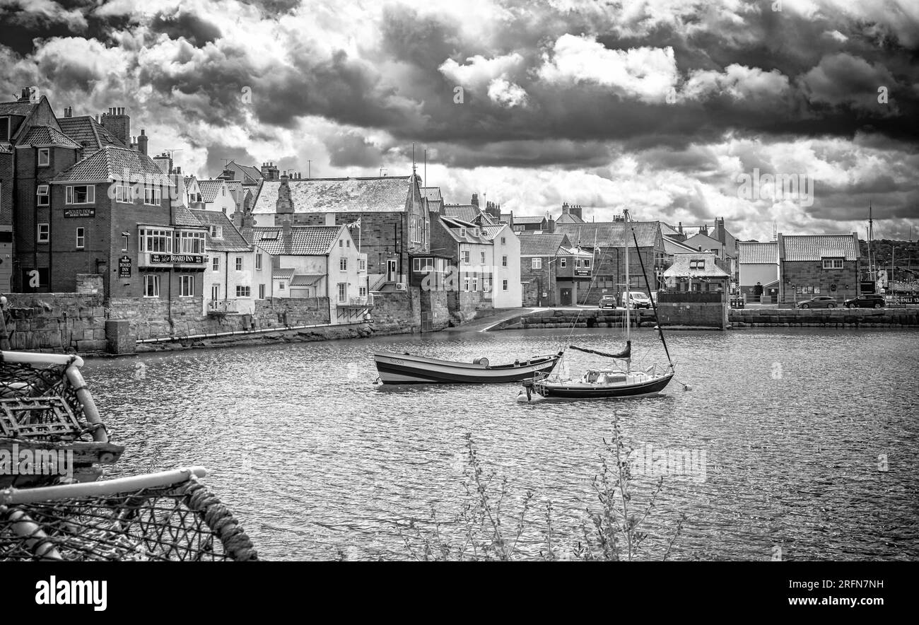 Two boats lie in harbour. Buildings line the waterfront and a quay is ...