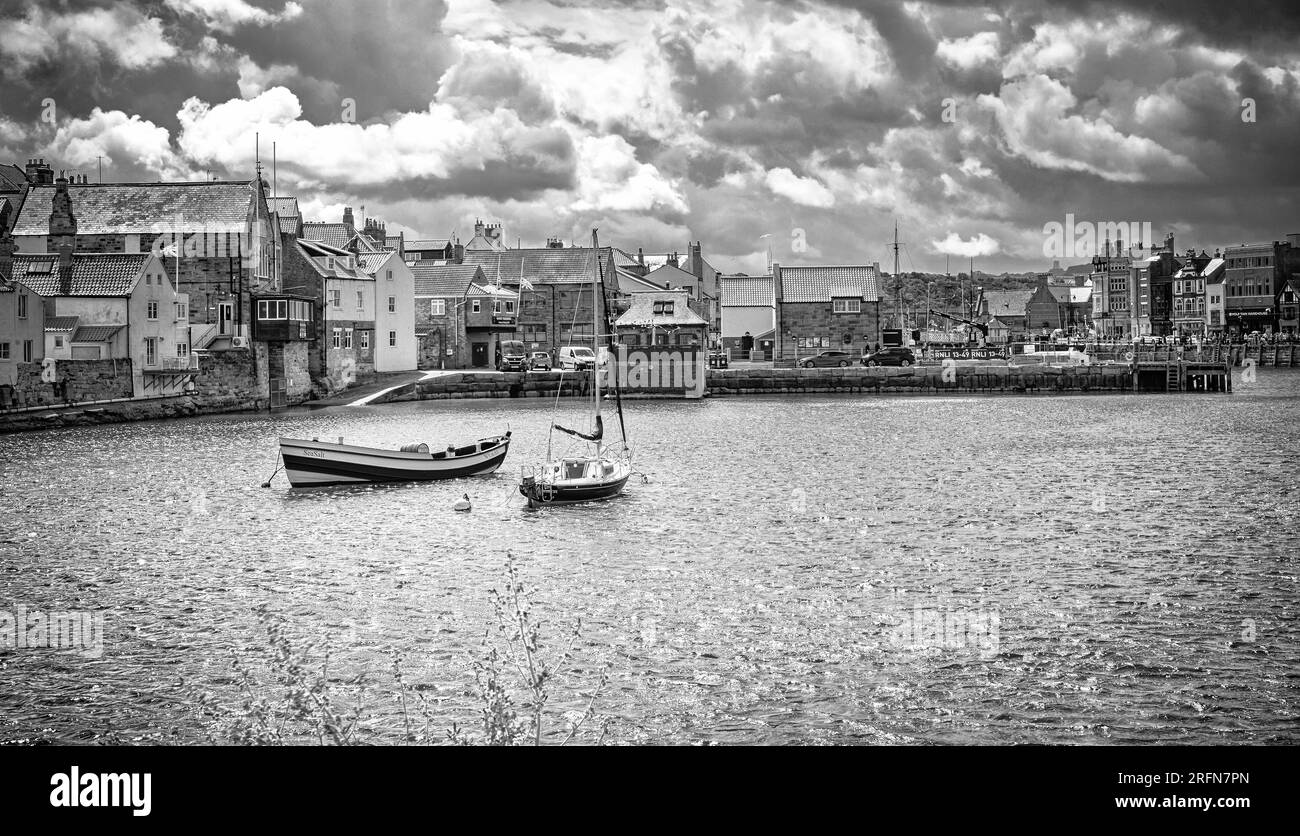 Two boats lie in harbour. Buildings line the waterfront and a quay is ...