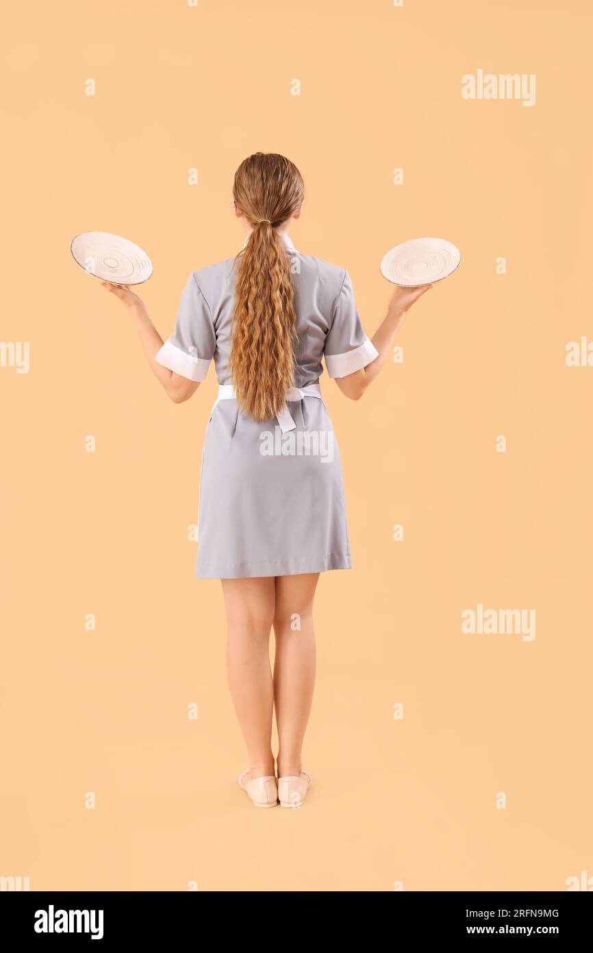 Beautiful young waitress with empty plates on beige background, back ...