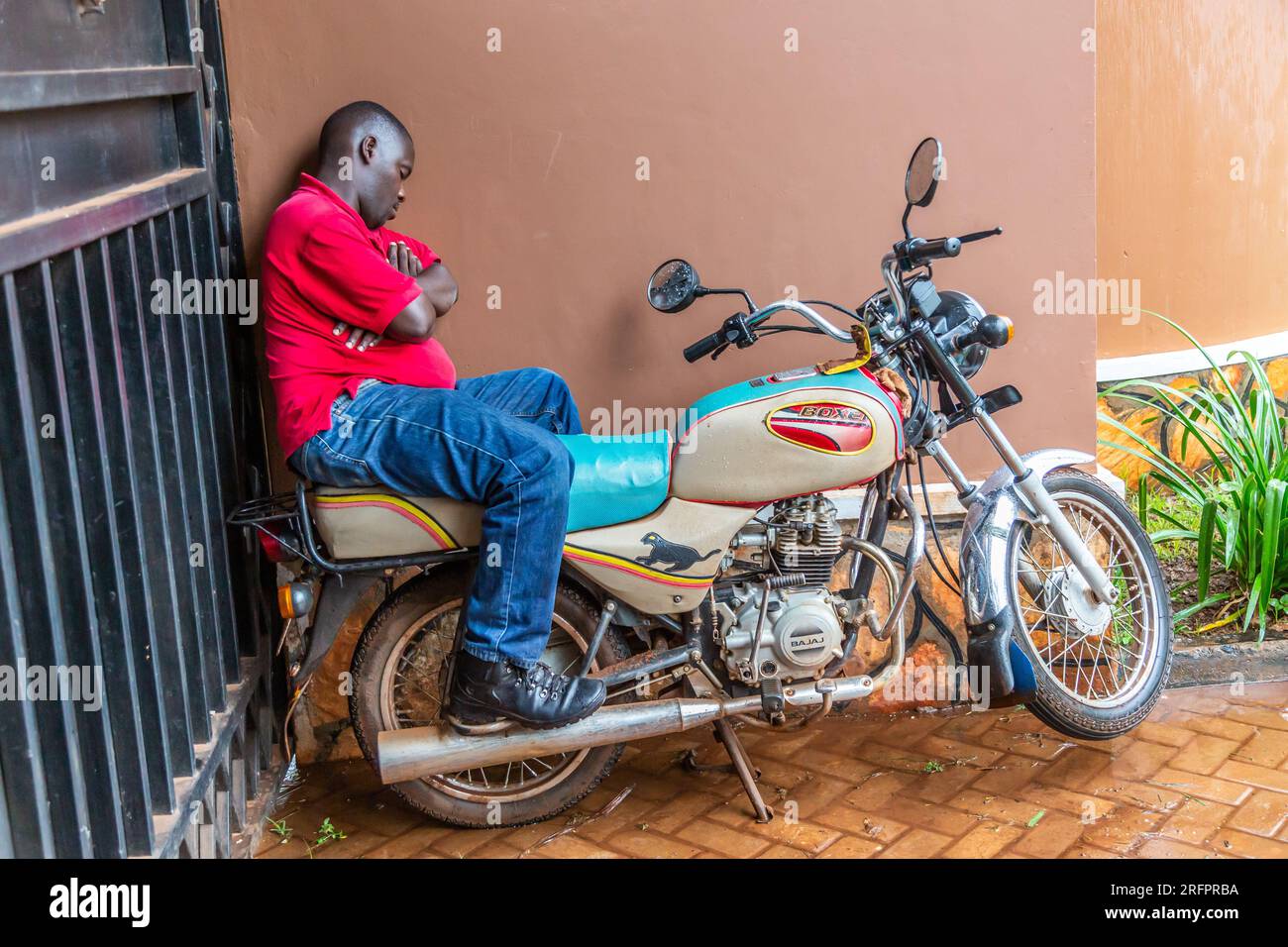 Sitting on his motorcycle, leaning against a wall, a boda-boda driver ...