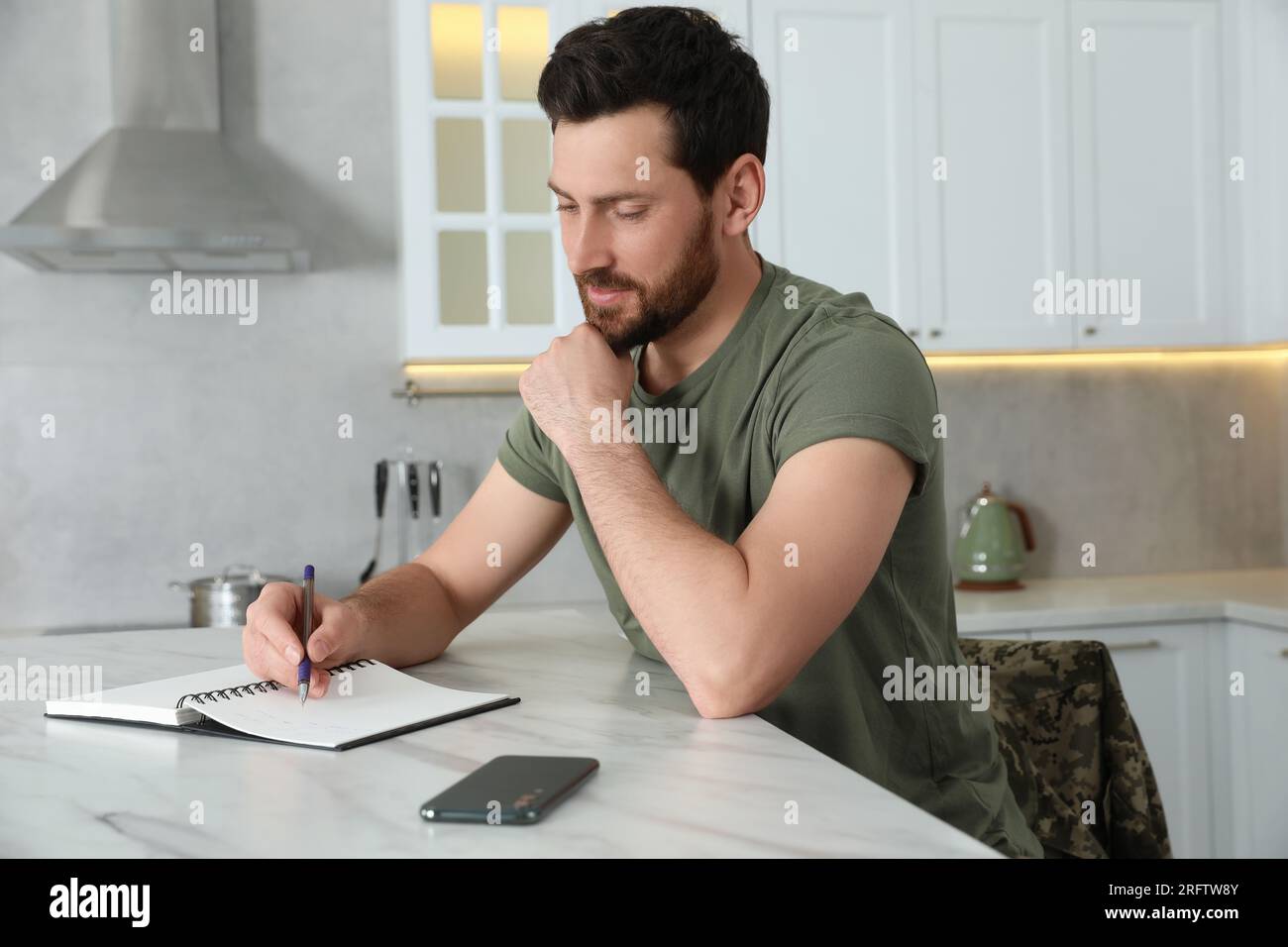 Soldier writing in notebook at white marble table in kitchen. Military ...
