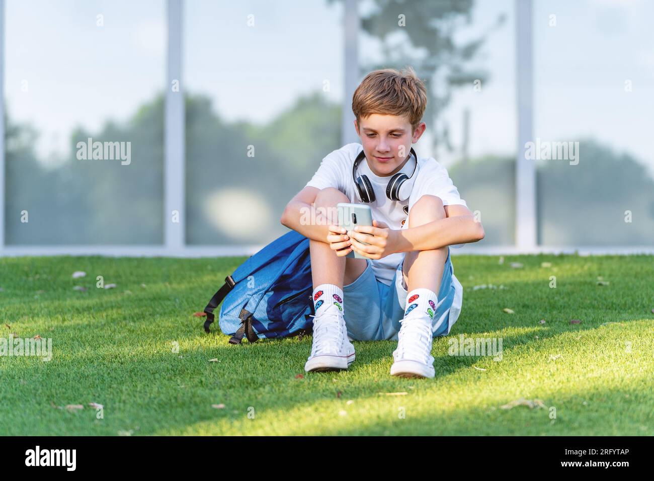 Teenage boy using his mobile phone while sitting on grass with backpack ...