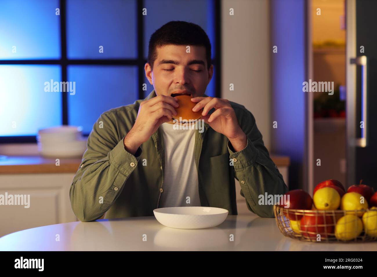 Hungry young man eating burger in kitchen at night Stock Photo - Alamy