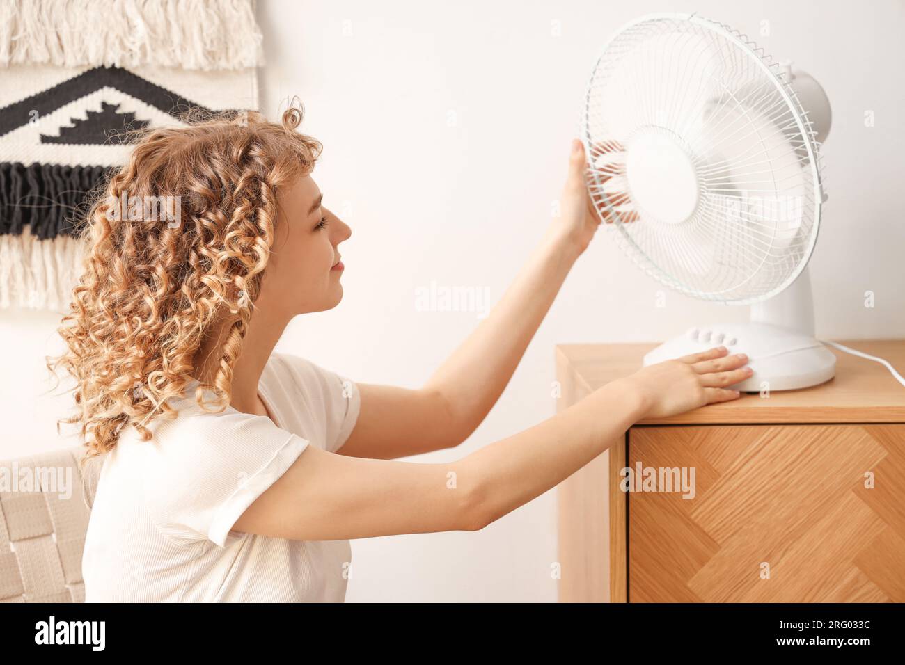 Beautiful happy young woman with electric fan sitting on chair at home ...