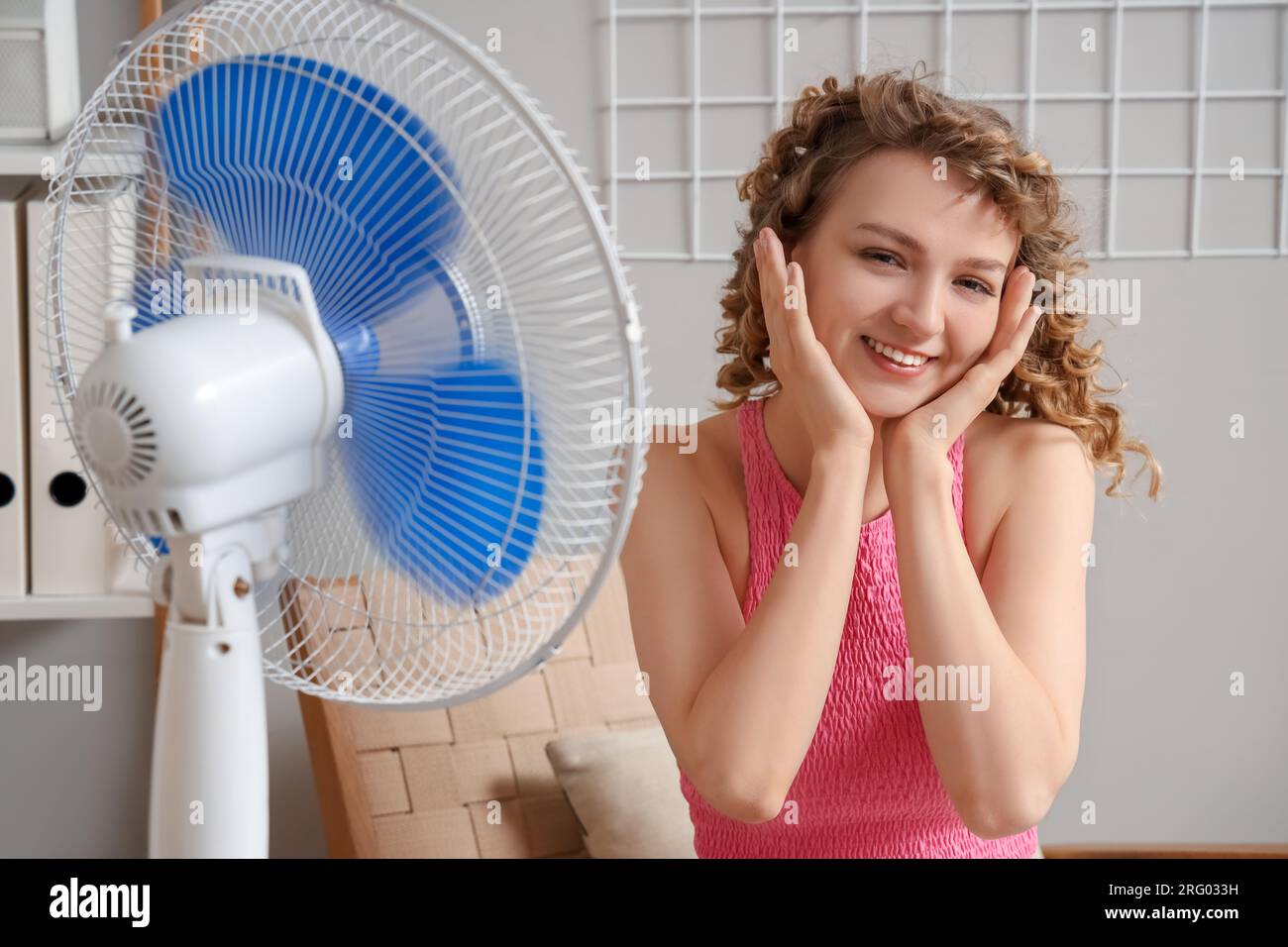 Beautiful happy young woman with electric fan sitting on chair in ...