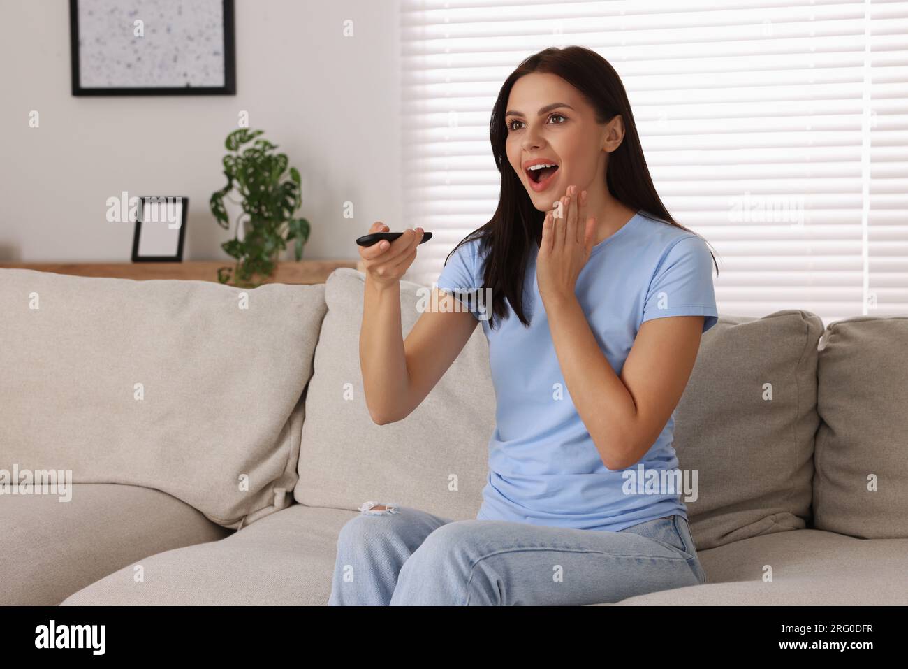 Emotional woman with remote controller watching TV on sofa at home ...