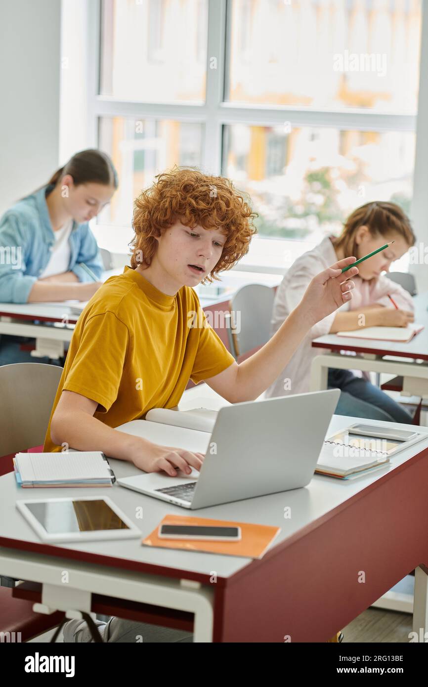 Redhead teen schoolboy holding pencil and using laptop while talking ...