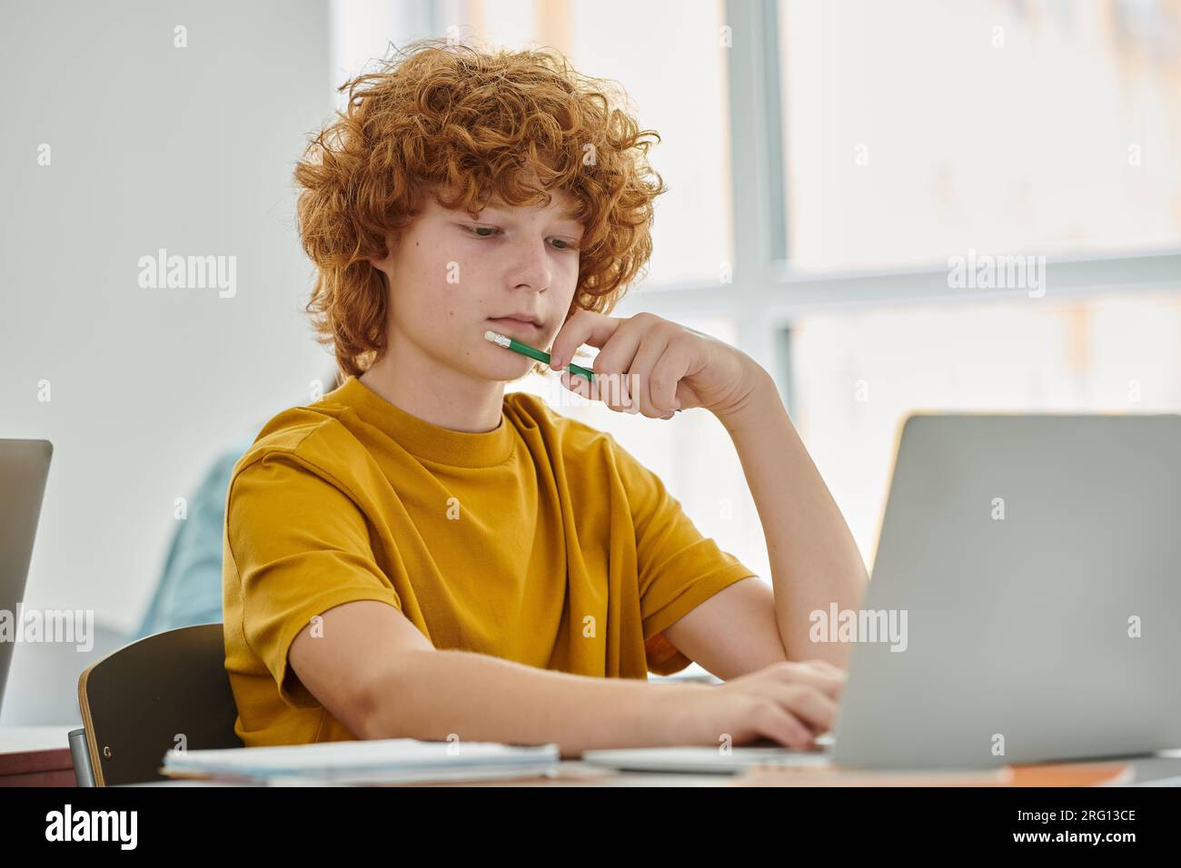 Redhead teenage schoolboy holding pencil and using laptop during lesson ...