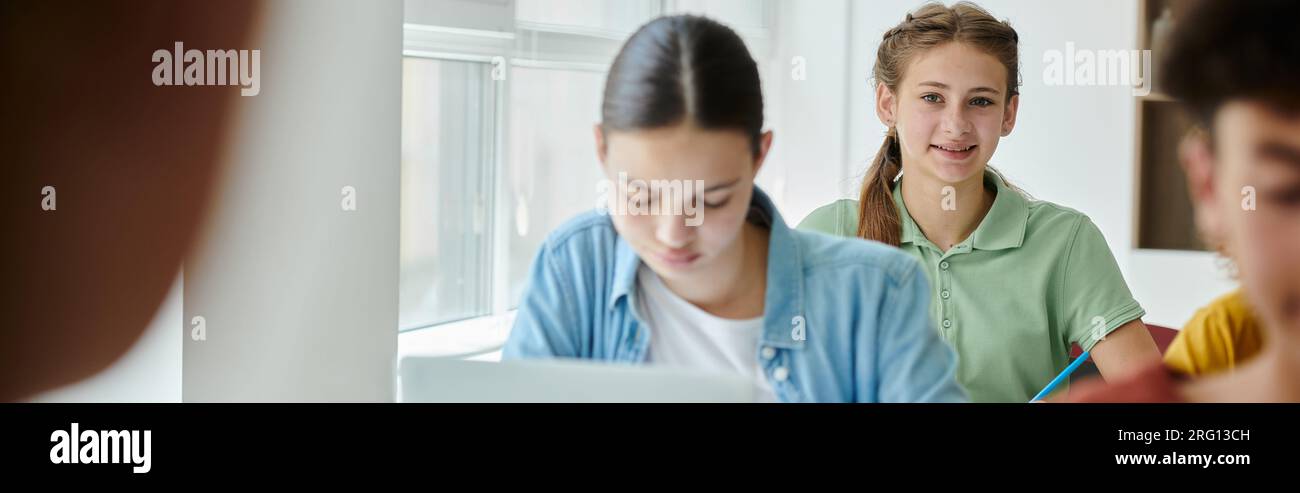 Smiling teenage schoolgirl looking at camera near blurred classmates ...