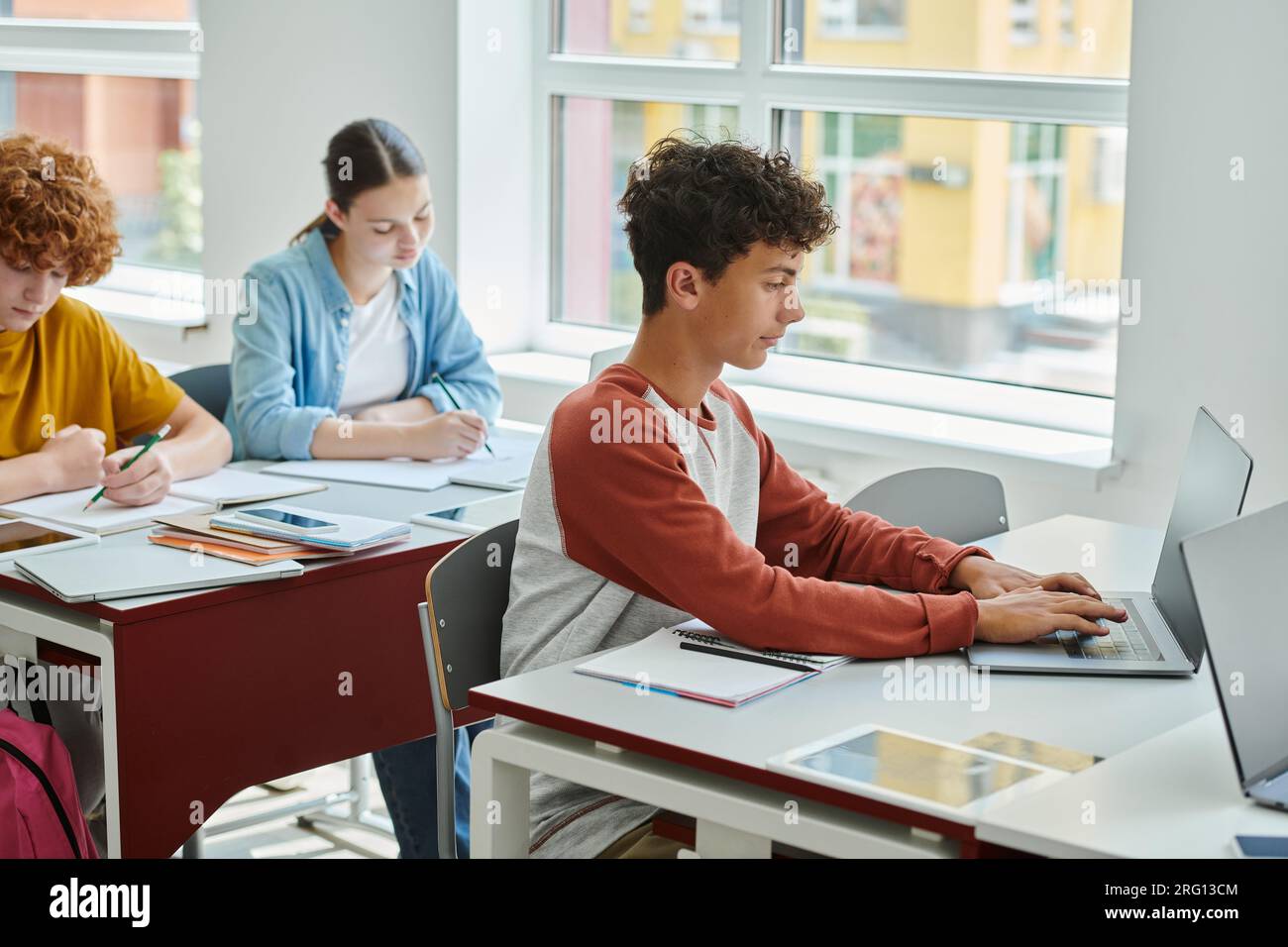 Teenage schoolboy using laptop near notebooks and blurred classmates in ...