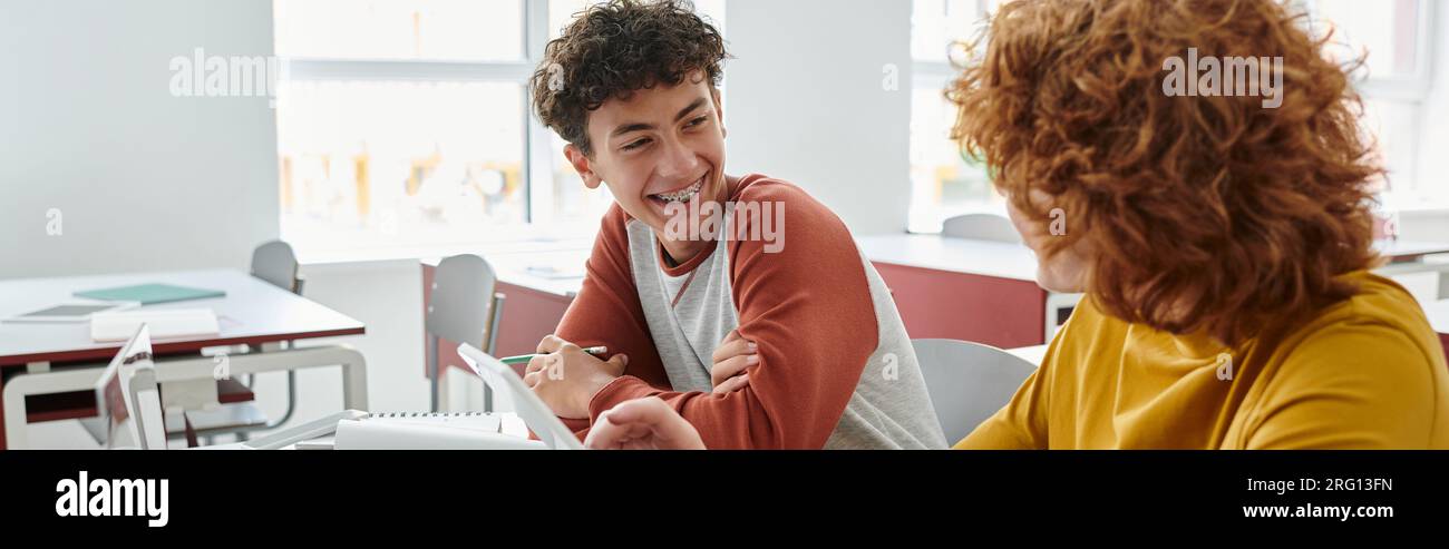 Smiling teen schoolboy talking to classmate near devices during lesson ...